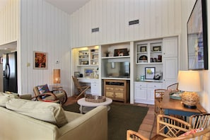 Uma sala de estar com sofá, cadeiras, televisão, mesa de jantar e área de cozinha com armários e geladeira.
