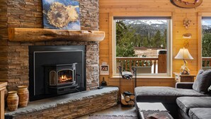 Interior acolhedor de uma cabana com lareira de pedra, paredes de madeira, um sofá confortável e vista para uma montanha nevada.