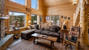 Interior acolhedor de uma cabana de toras, com um amplo sofá modular, uma poltrona de balanço de madeira e uma lareira.