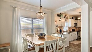 Uma sala de jantar com mesa de madeira e cadeiras brancas, um candelabro e vista para a cozinha através de uma janela com persianas.