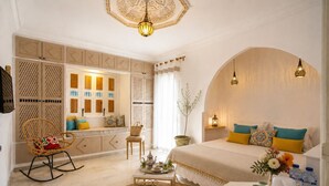 Quarto com cama, área de estar com almofadas, mesa com bule e luminária decorativa no teto.