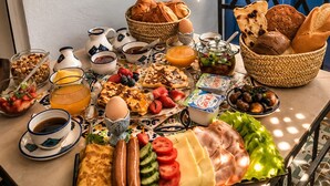 Mesa de café da manhã com diversos alimentos, uma cesta de palha e uma porta azul ao fundo.
