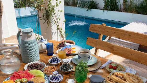 Mesa de café da manhã com diversas comidas, um bule e vista para uma piscina.