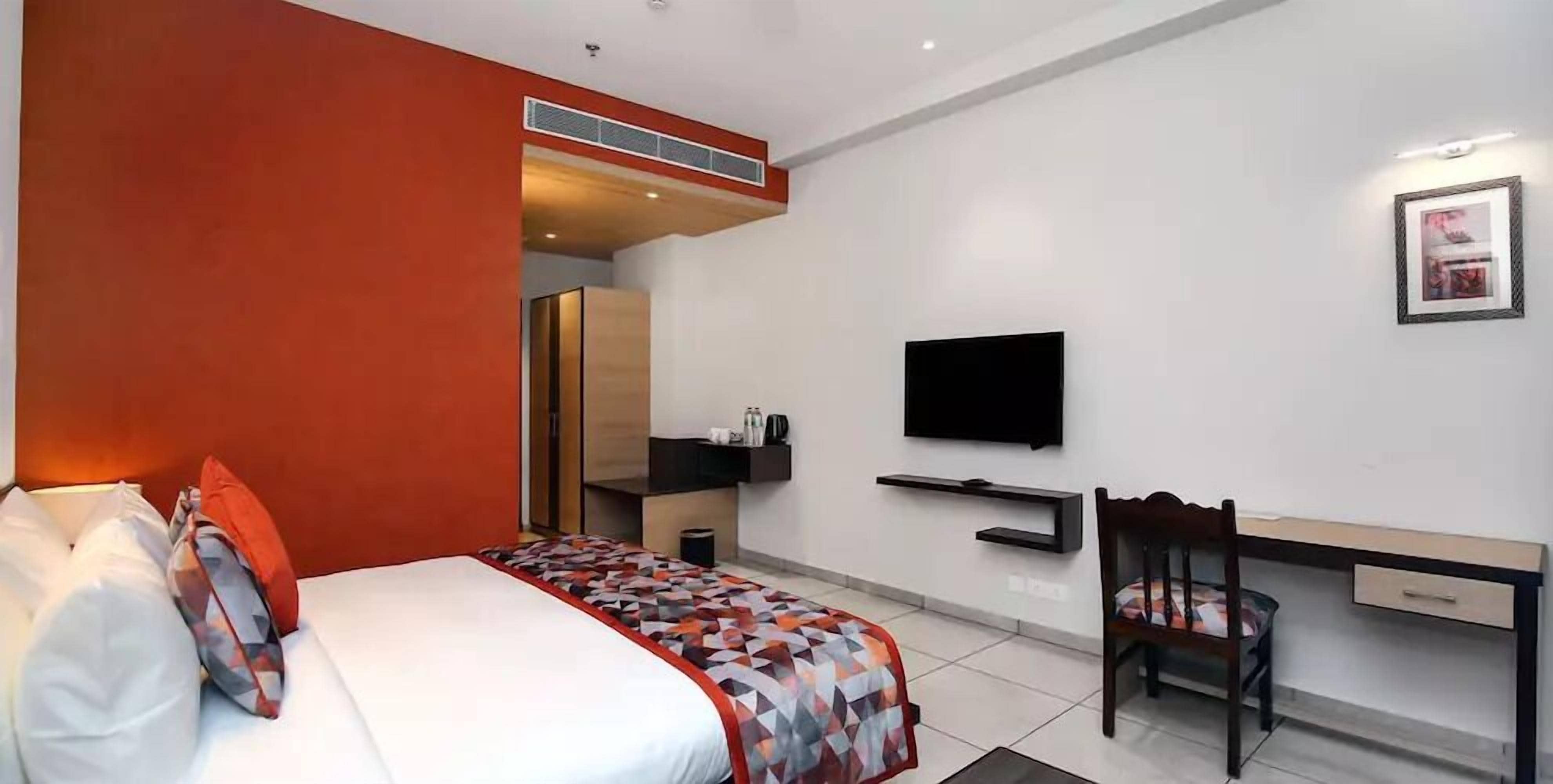 Quarto de hotel com cama, escrivaninha, cadeira, televisão e guarda-roupa.