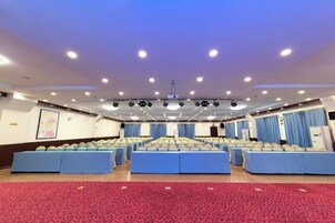 Uma sala de conferências com fileiras de assentos azuis, um tapete vermelho e um palco com um projetor.