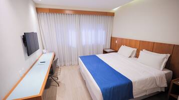 Quarto de hotel com cama, escrivaninha, televisão e janela com cortinas.