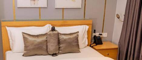 Cama bem arrumada com cabeceira marrom, um travesseiro e uma toalha dobrada. Um criado-mudo com um telefone e um abajur. Duas fotos emolduradas penduradas na parede acima da cama.
