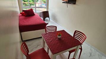 Quarto com cama, mesa e cadeiras, televisão na parede e janela com vista para árvores.