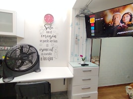 Quarto com escrivaninha, relógio de parede, televisão de tela plana, ventilador e parede decorativa com uma citação.