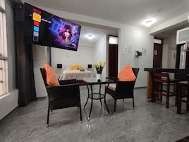 Quarto de hotel com cama, televisão na parede, mesa de jantar com cadeiras e uma pequena mesa com um vaso de flores.
