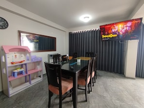 Uma área de refeições com uma mesa de vidro, cadeiras de madeira, uma casinha de brinquedo rosa e uma TV exibindo um programa.