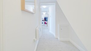 Um corredor estreito com paredes brancas e chão coberto de carpete, que leva a um quarto com área de lavanderia visível ao fundo.