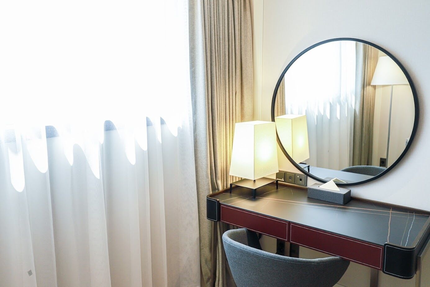 Quarto de hotel com mesa de toucador, espelho, duas luminárias e uma cadeira.