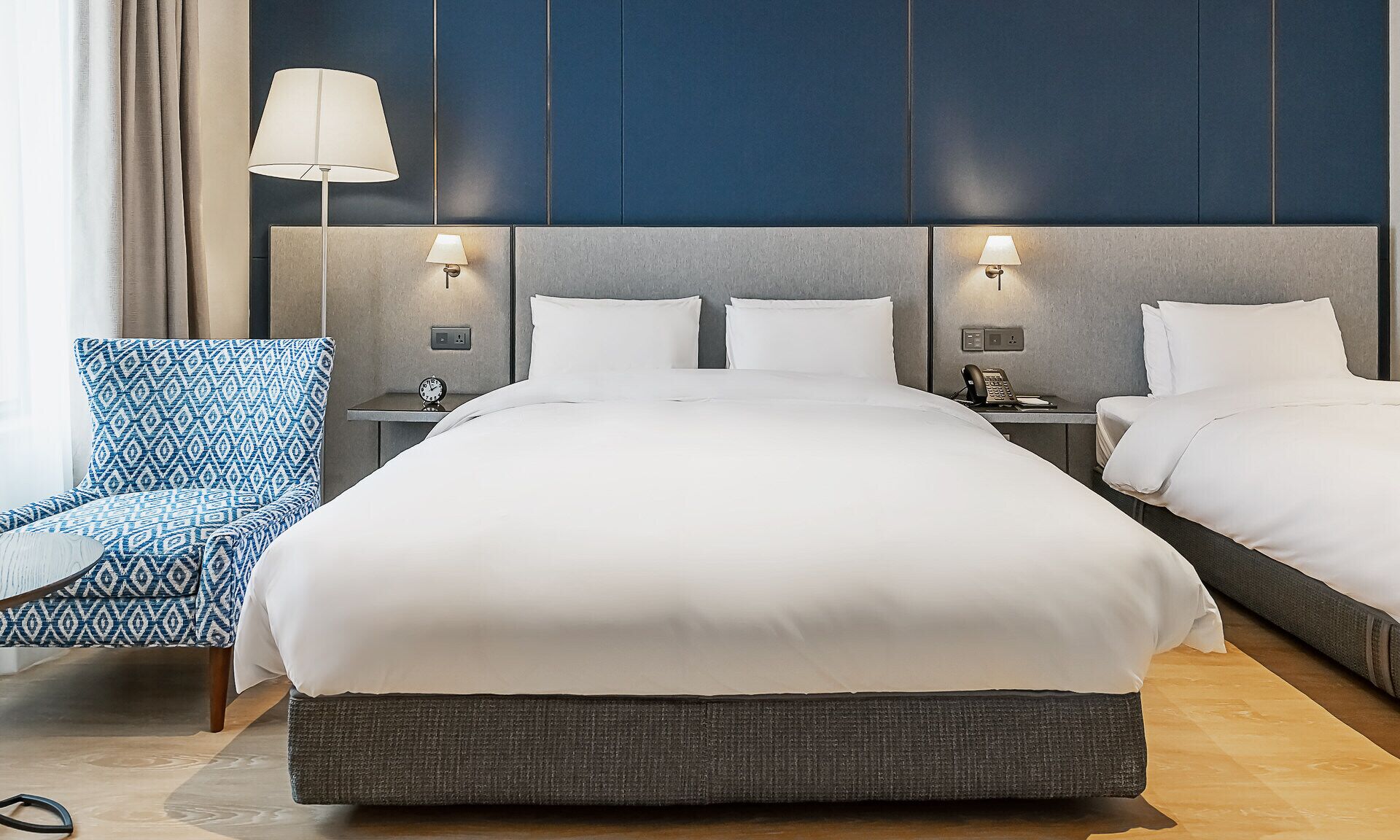 Quarto de hotel com uma cama grande, duas mesas de cabeceira com luminárias, uma poltrona estampada e um telefone fixado na parede.