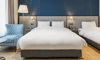 Quarto de hotel com uma cama grande, duas mesas de cabeceira com luminárias, uma poltrona estampada e um telefone fixado na parede.