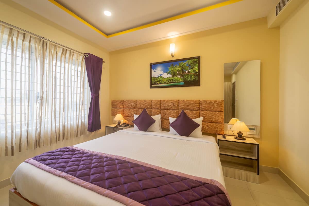 Quarto de hotel com uma cama, mesa de cabeceira, luminária, quadro e janela com cortinas.