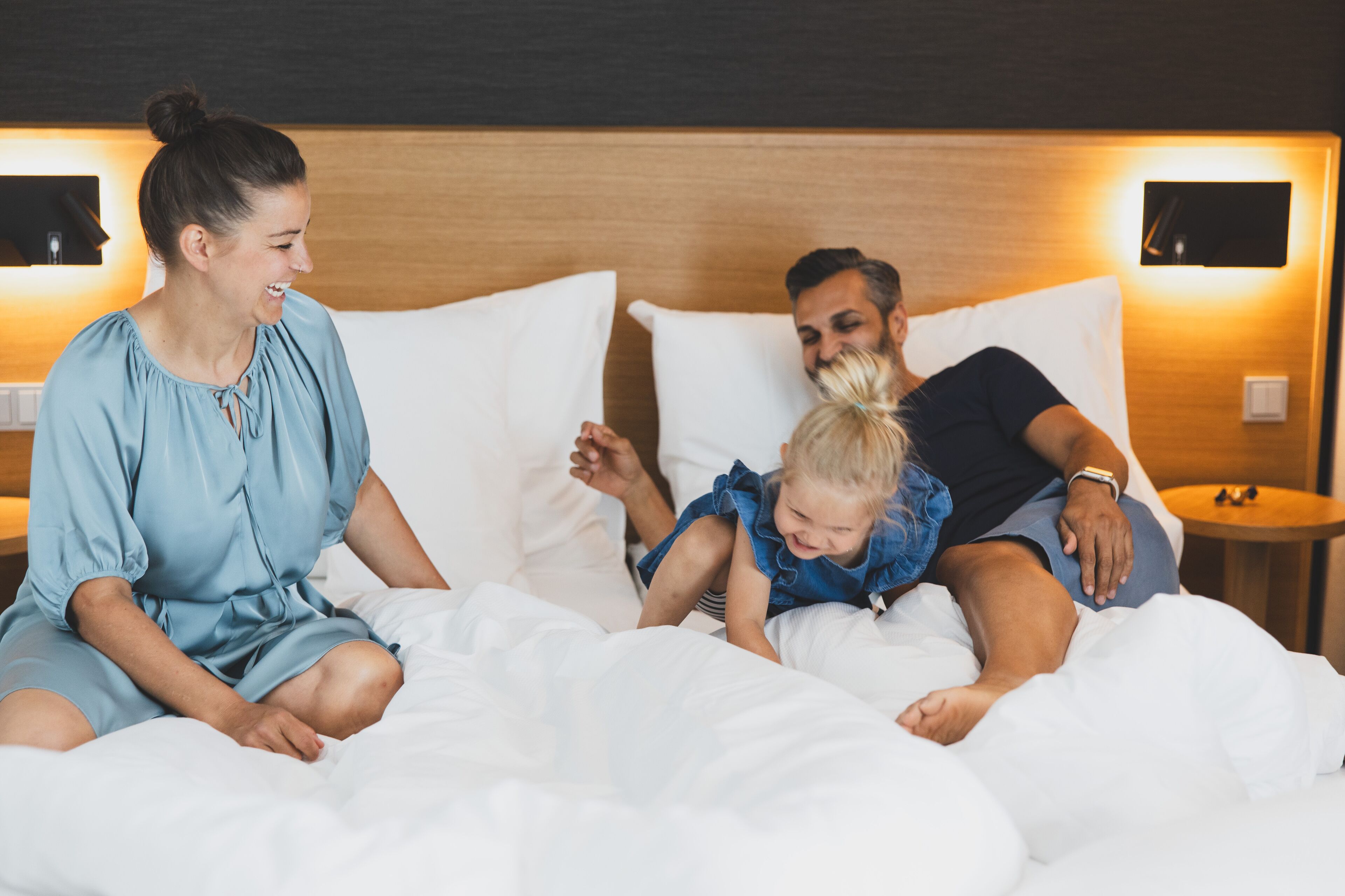Uma família de três pessoas em quarto de hotel, com cabeceira de madeira, roupa de cama branca e mesas de cabeceira com abajures.