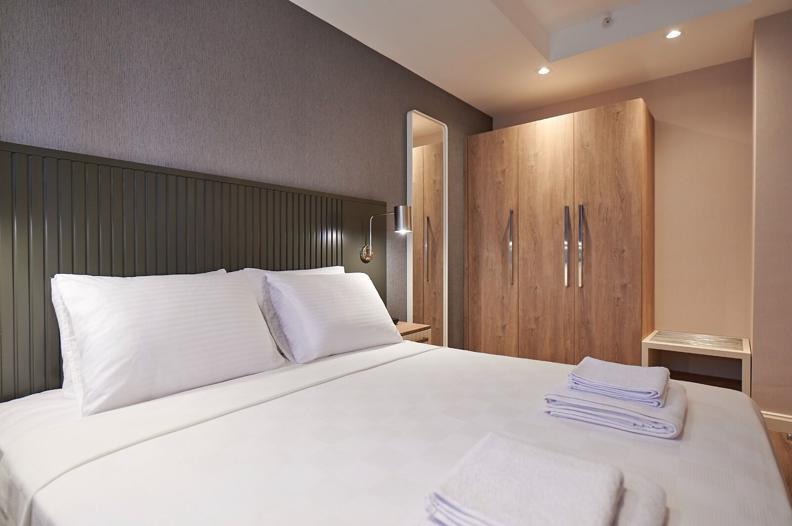 Quarto de hotel moderno com uma cama grande, um guarda-roupa de madeira e uma mesinha de cabeceira.