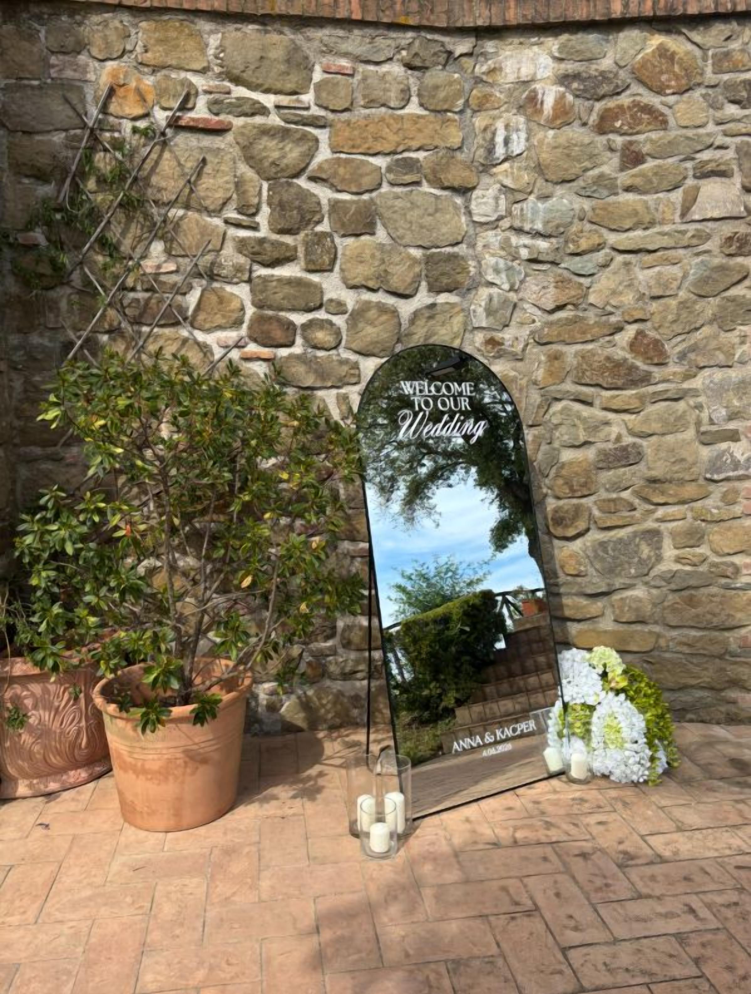 Mural de pedra com placa de boas-vindas, um vaso de planta e um pequeno arranjo floral.