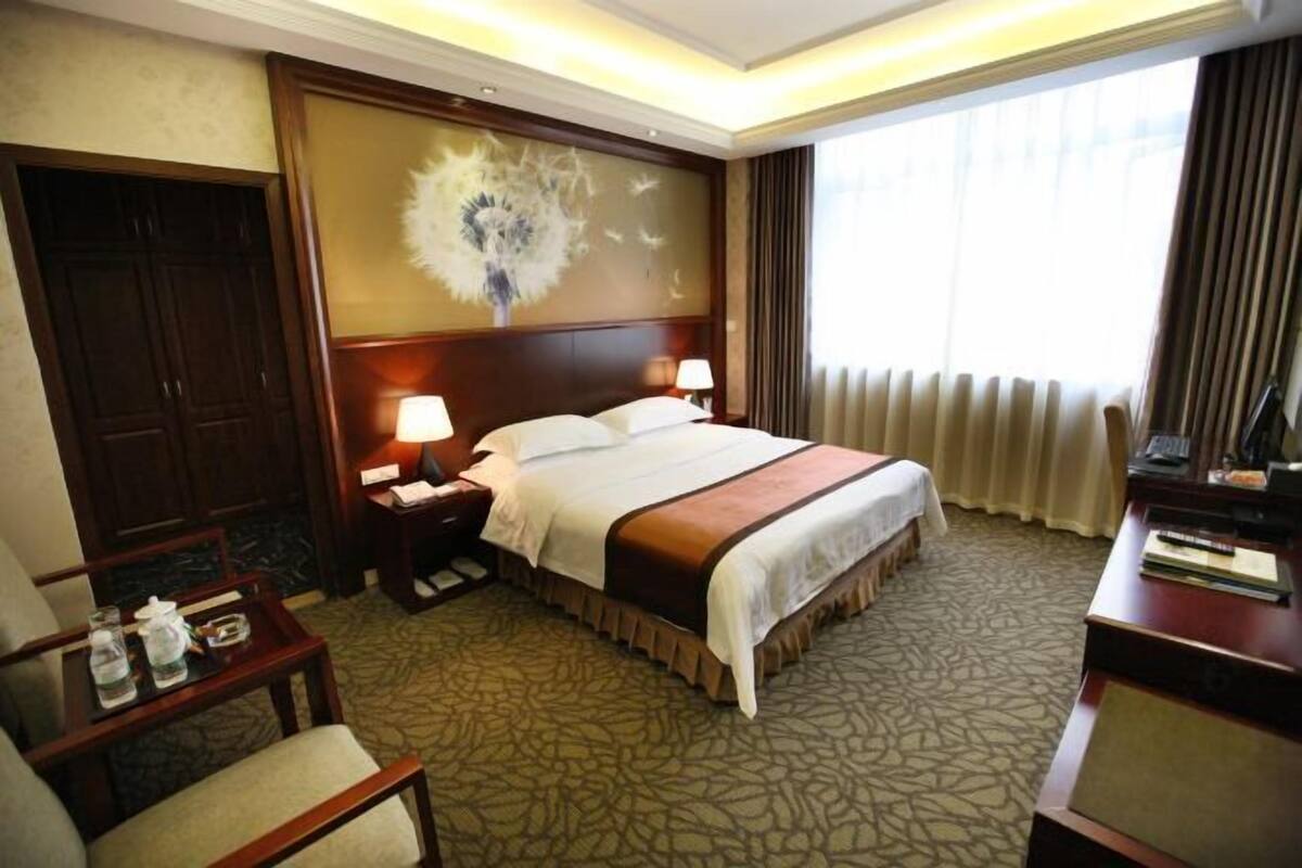 Quarto de hotel com uma cama grande, duas mesas de cabeceira com luminárias, uma escrivaninha, uma cadeira, um guarda-roupa e uma janela grande com cortinas.