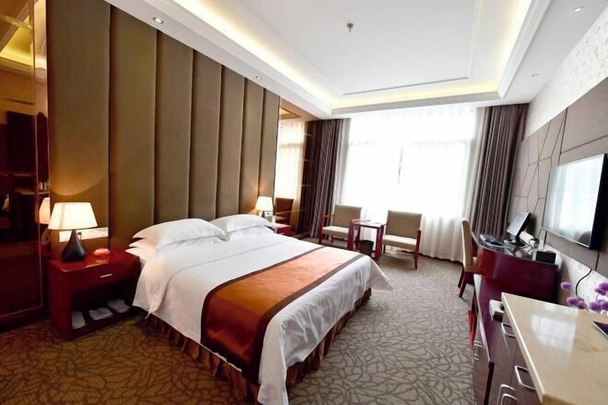 Quarto de hotel com cama grande, escrivaninha, cadeira, televisão e janela com cortinas.