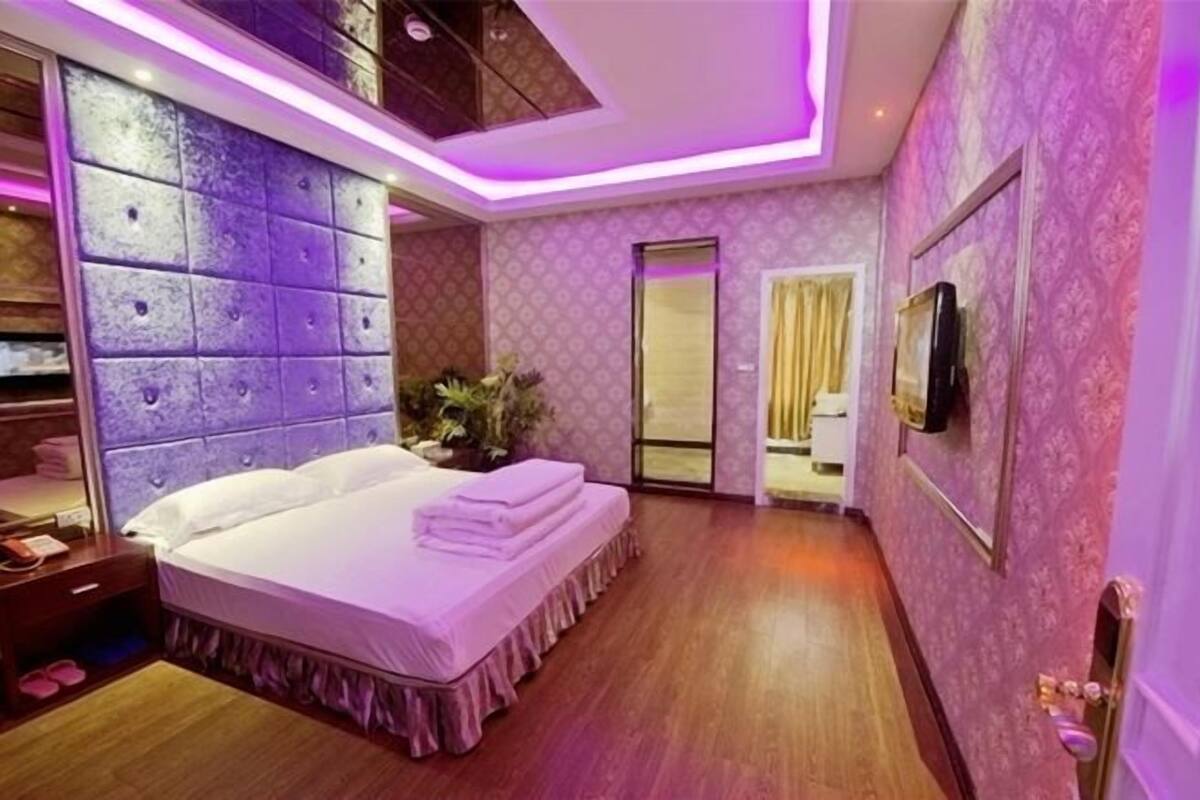 Quarto moderno com cama grande, TV fixada na parede e iluminação roxa no teto.