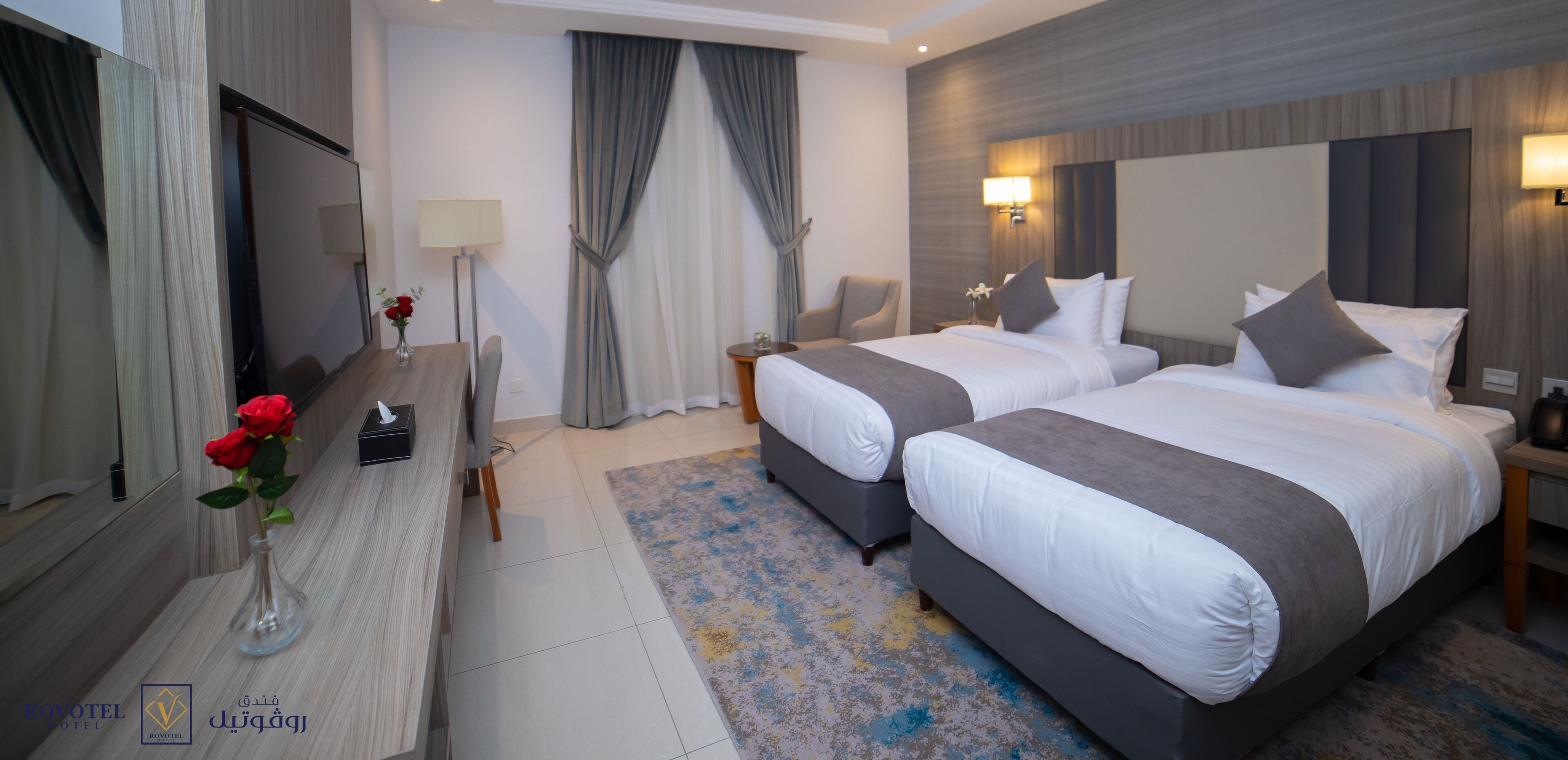 Quarto de hotel com duas camas, uma secretária com um vaso de flores, uma luminária e um espelho grande.