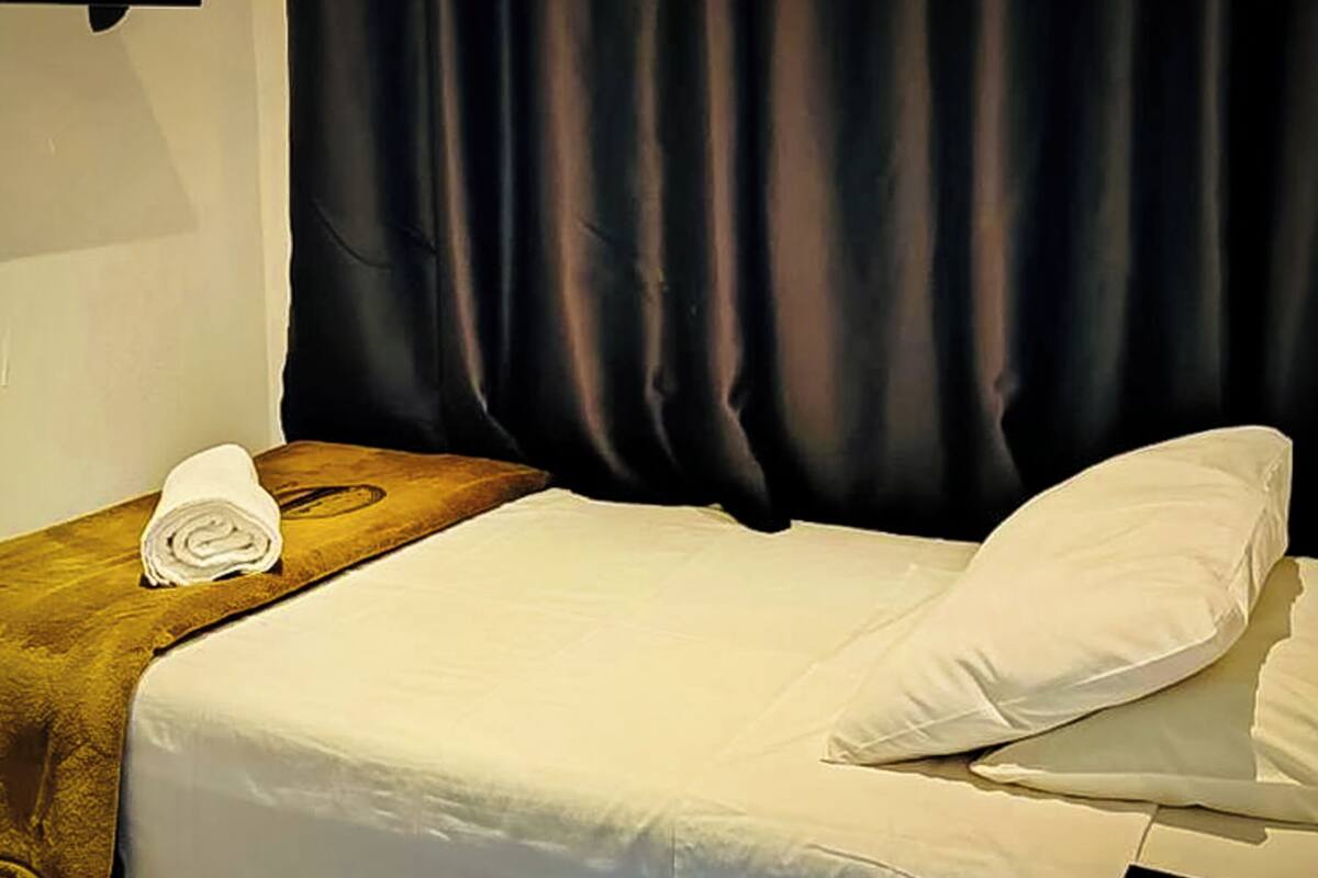 Quarto de hotel com uma cama bem arrumada, cabeceira de madeira e cortina preta.
