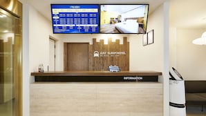 Balcão de recepção de hotel com um visor digital acima, área de recepção em madeira e vista para um quarto.