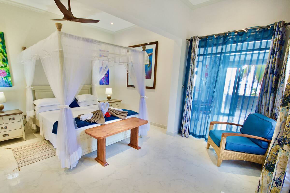 Quarto com cama de dossel, mesa de cabeceira com abajur, um banquinho, cadeira de vime e uma janela grande com cortinas.