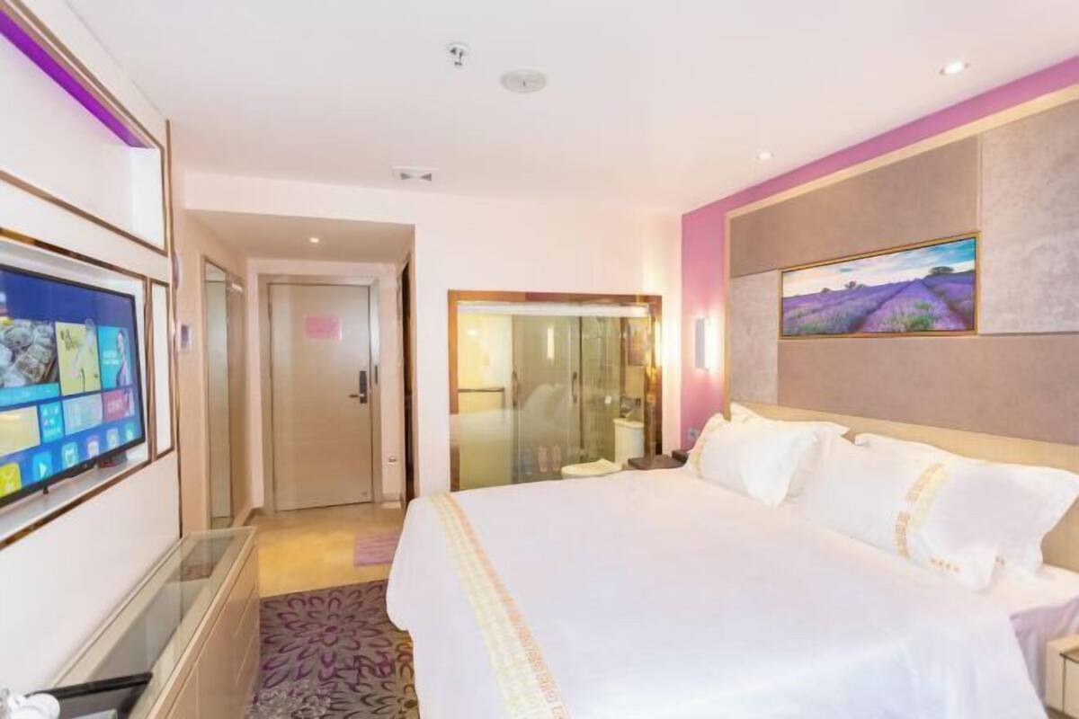Quarto de hotel com uma cama grande, televisão na parede, box de chuveiro de vidro e um quadro na parede.