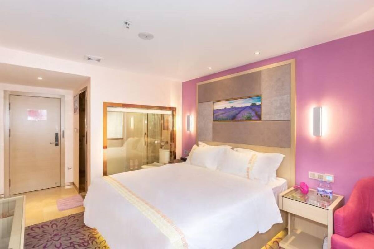 Quarto de hotel com uma cama grande, porta de vidro que dá para um banheiro e um quadro na parede.