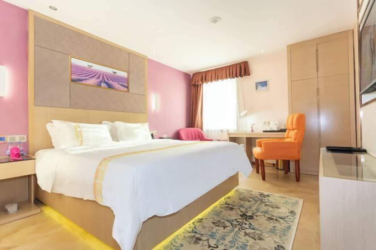 Quarto de hotel com cama grande, escrivaninha, poltrona laranja, televisão e janela com cortinas.