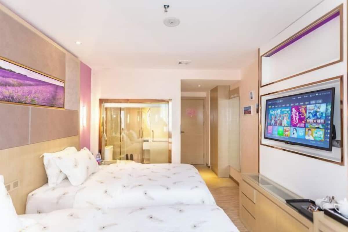 Quarto de hotel com uma cama, uma televisão, um quadro e uma porta de vidro que dá para um banheiro.