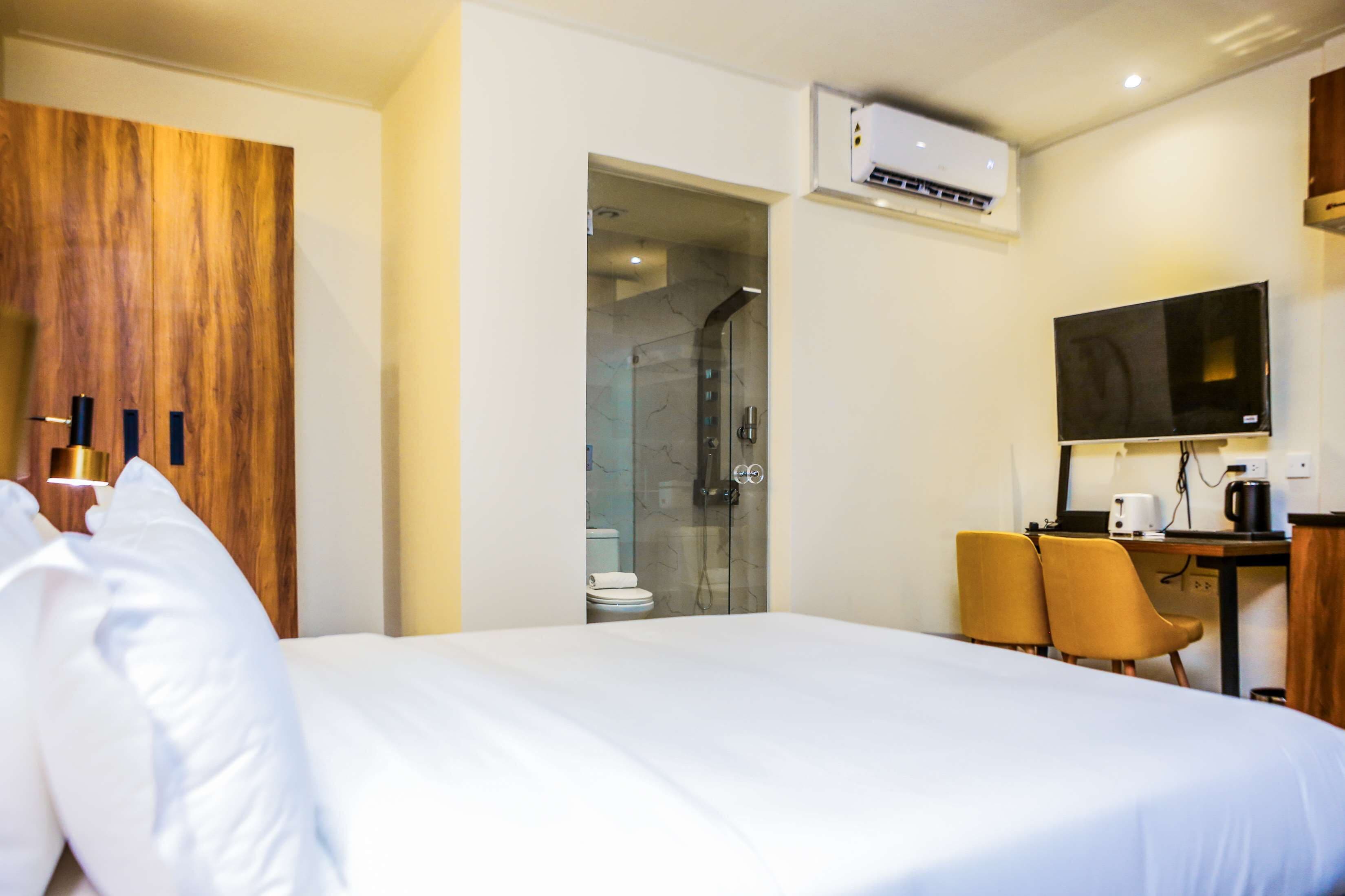 Quarto de hotel com uma cama grande, uma escrivaninha com duas cadeiras, uma televisão e um ar condicionado.