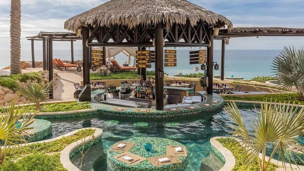 Uma área de piscina em resort, com um gazebo de palha, espreguiçadeiras e um bar.