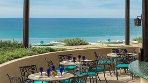 Terraço com mesas e cadeiras, vista para uma praia e área de piscina.