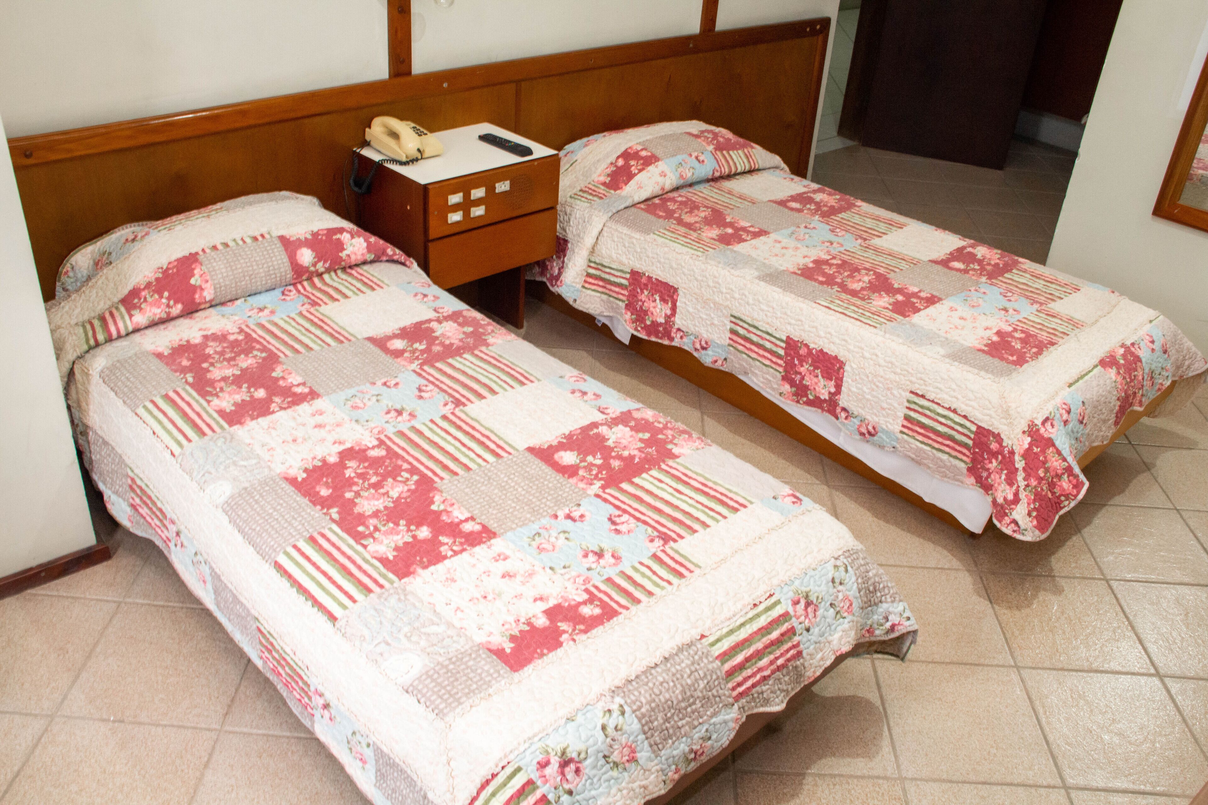 Duas camas de solteiro com colchas de padrão floral, uma cabeceira de madeira, uma mesa de cabeceira com telefone e piso de azulejos.