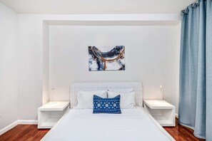 Quarto moderno com cama branca, duas mesas de cabeceira, um quadro abstrato azul e cortina azul.