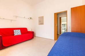 Um quarto com um sofá vermelho, uma cama azul, uma porta de madeira e um pequeno espelho na parede.