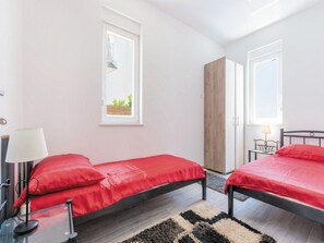 Quarto com duas camas de solteiro, guarda-roupa de madeira, mesa pequena com abajur e janela com vista para vegetação.