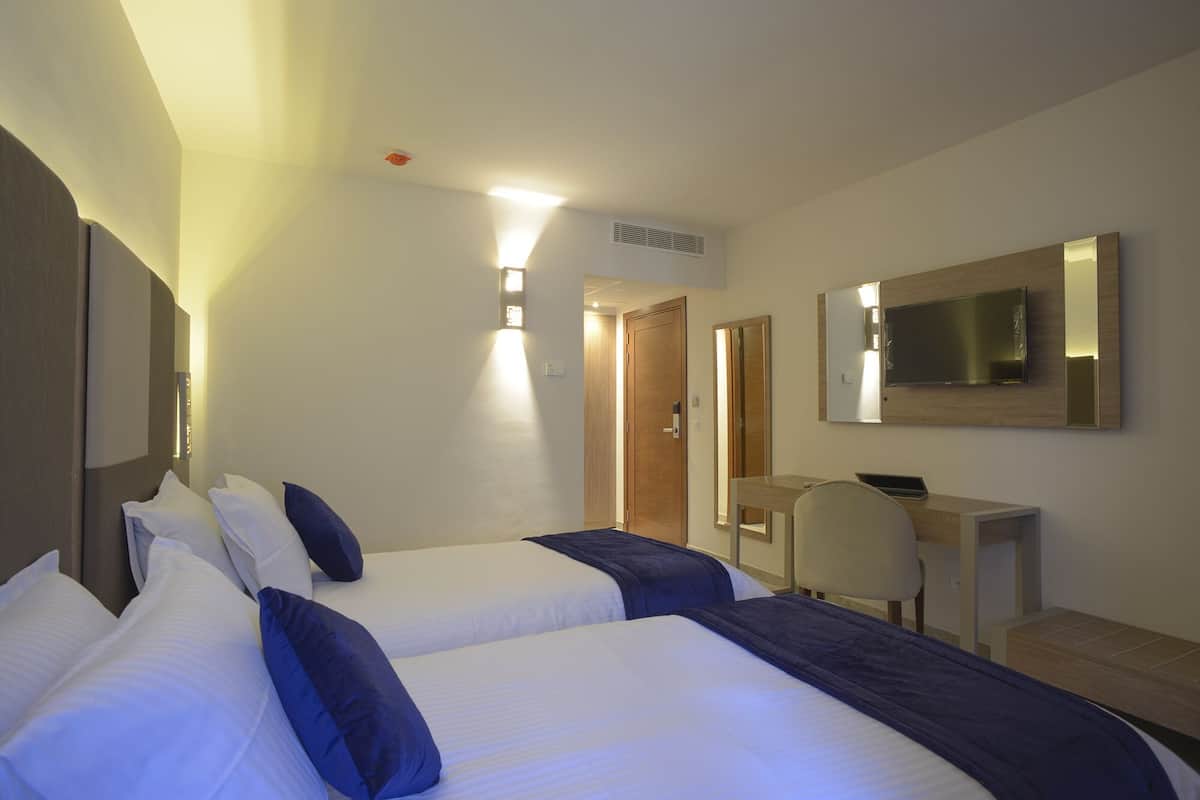 Quarto de hotel com duas camas, uma escrivaninha, uma cadeira e uma TV fixada na parede.