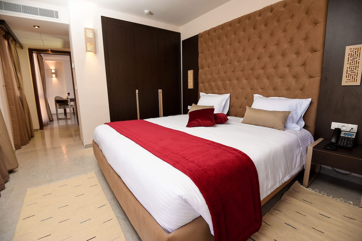 Quarto de hotel com uma cama grande, manta vermelha, mesa de cabeceira de madeira com telefone e guarda-roupa de madeira escura.