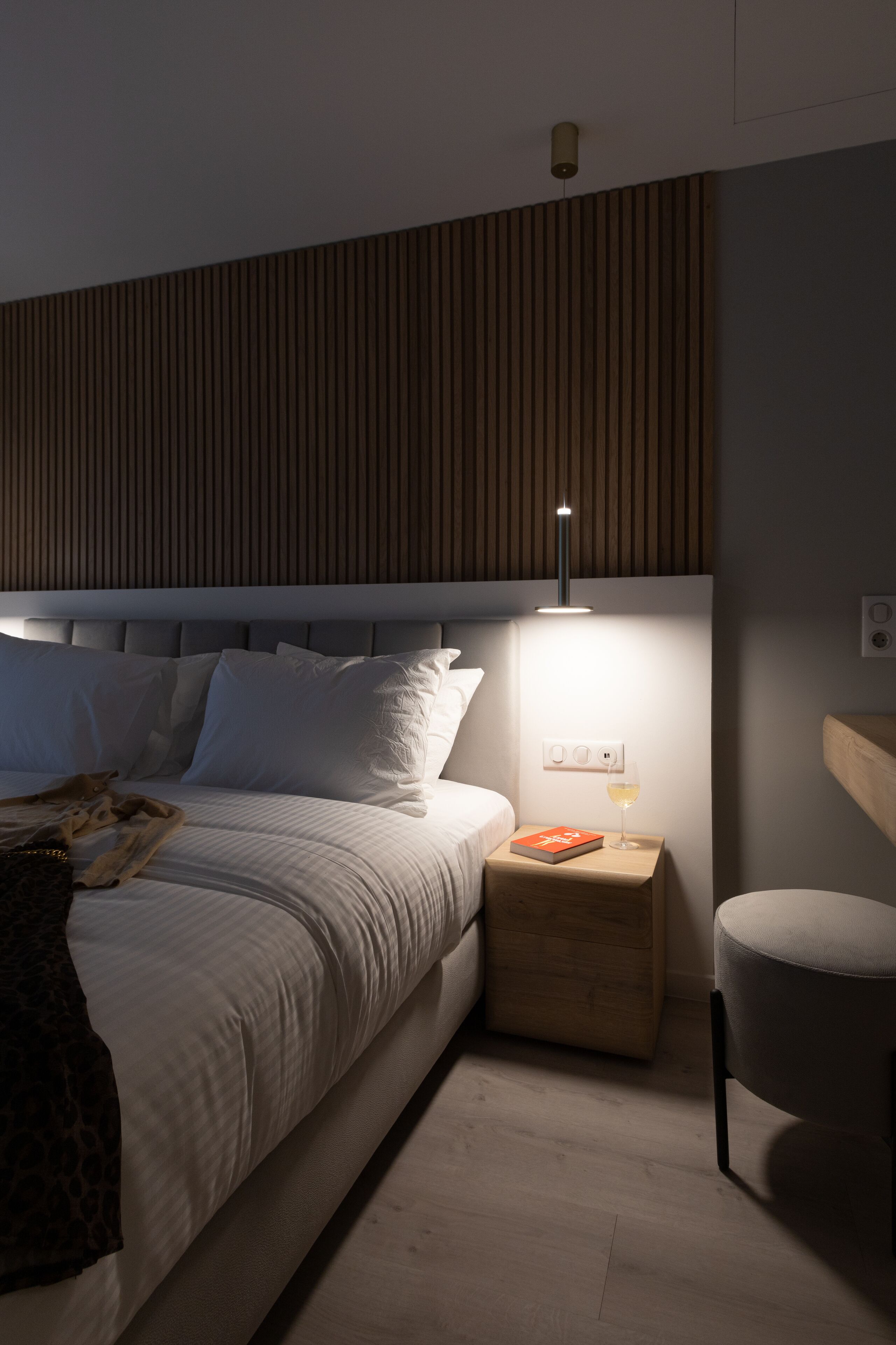 Quarto moderno com cama, mesa de cabeceira, abajur e um banquinho.