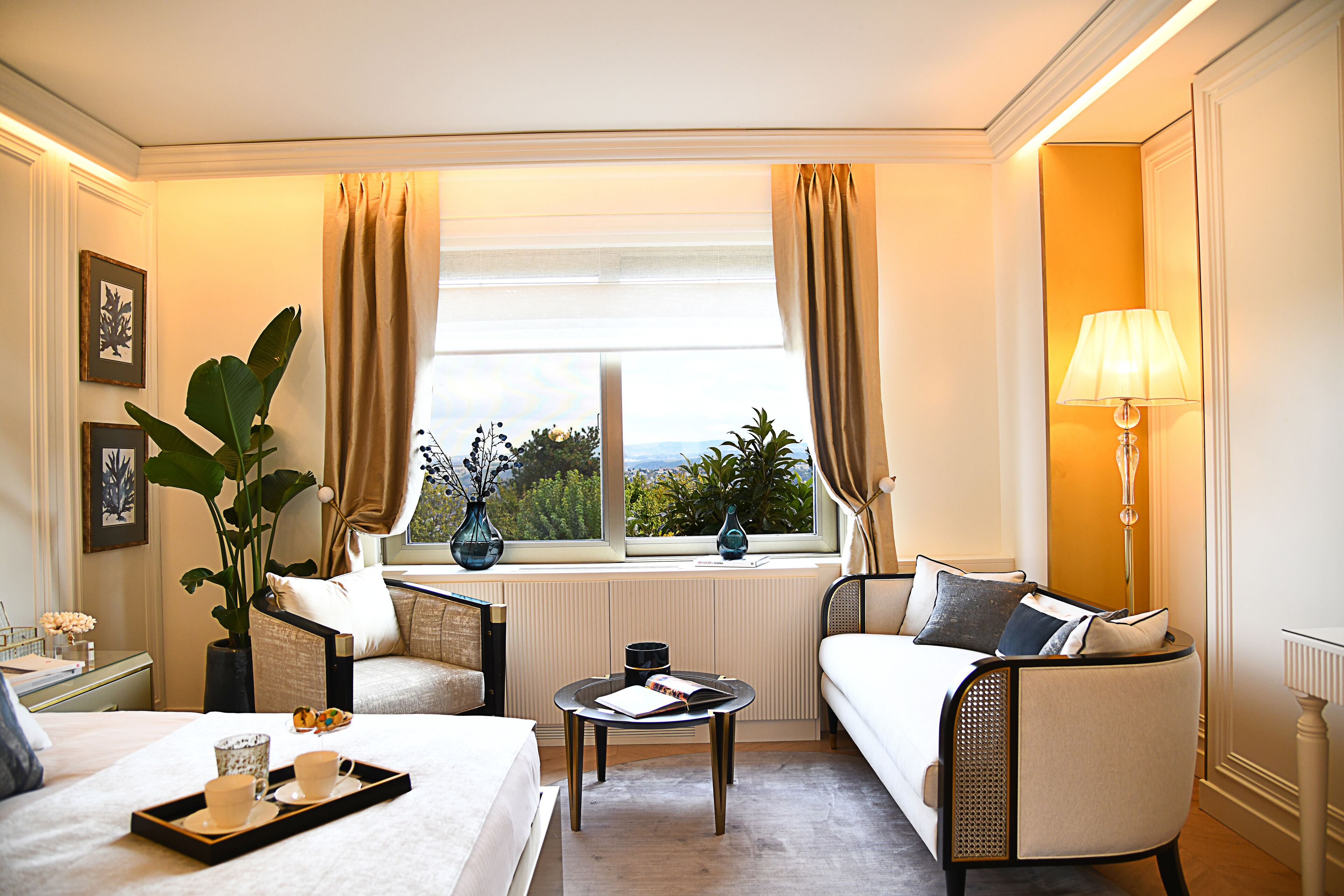 Quarto de hotel com cama, duas poltronas, uma mesinha, janela com cortinas, luminária e vista para o exterior.