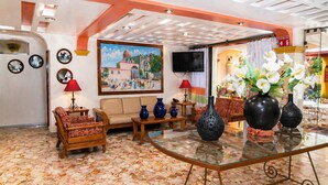 Um saguão de hotel com piso de azulejos, um conjunto de sofás, uma mesa de centro de vidro, vasos decorativos e um quadro na parede.