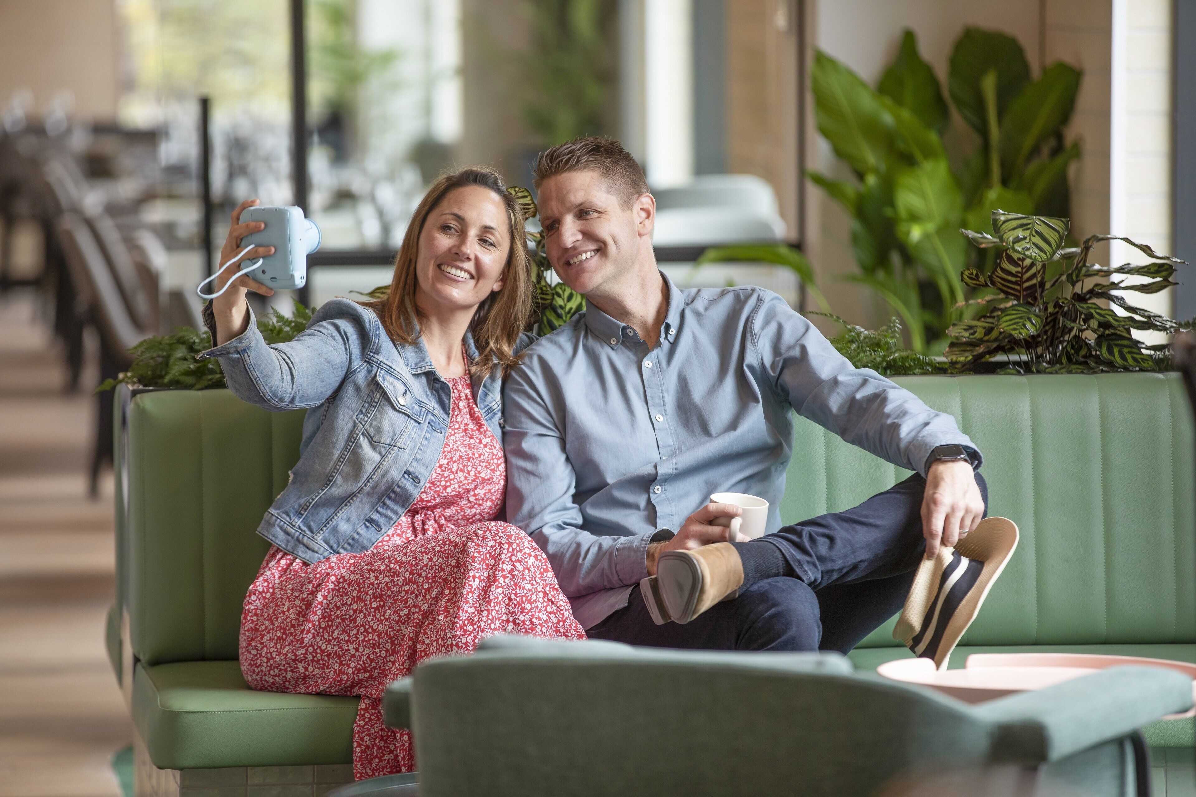 Um casal sentado em um sofá verde em ambiente interno bem iluminado, com plantas e vista para uma área de jantar ao fundo.