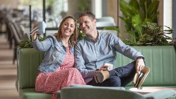 Um casal sentado em um sofá verde em ambiente interno bem iluminado, com plantas e vista para uma área de jantar ao fundo.