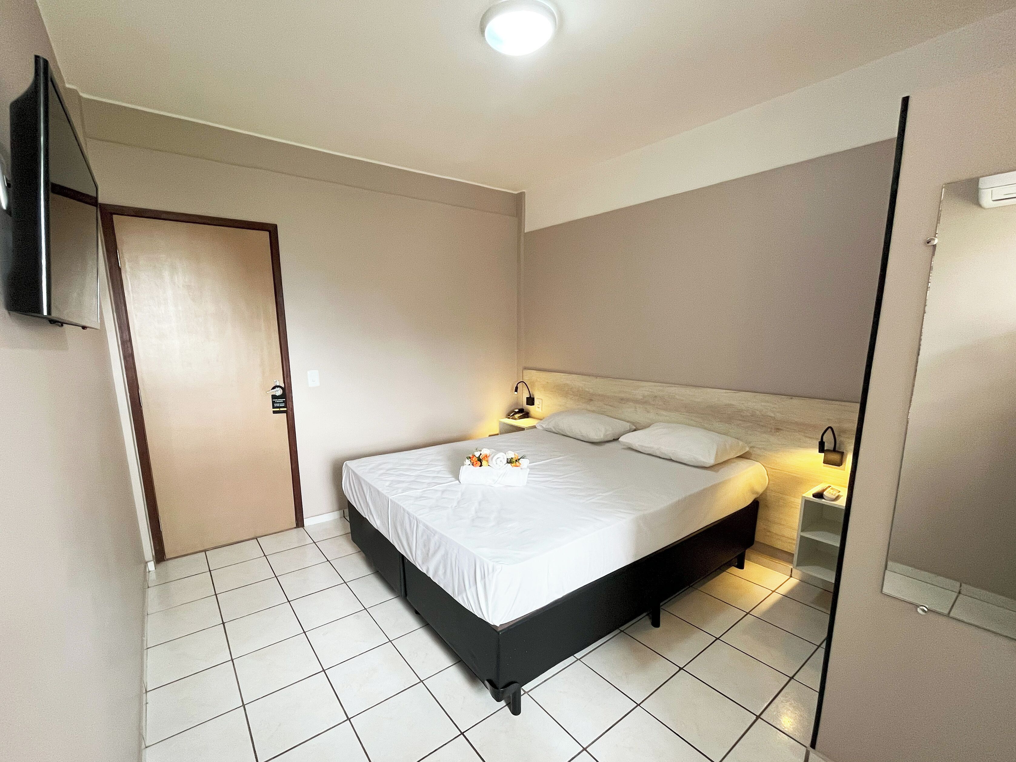 Quarto de hotel com cama, mesa de cabeceira, luminária e televisão na parede.