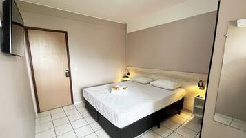 Quarto de hotel com cama, mesa de cabeceira, luminária e televisão na parede.